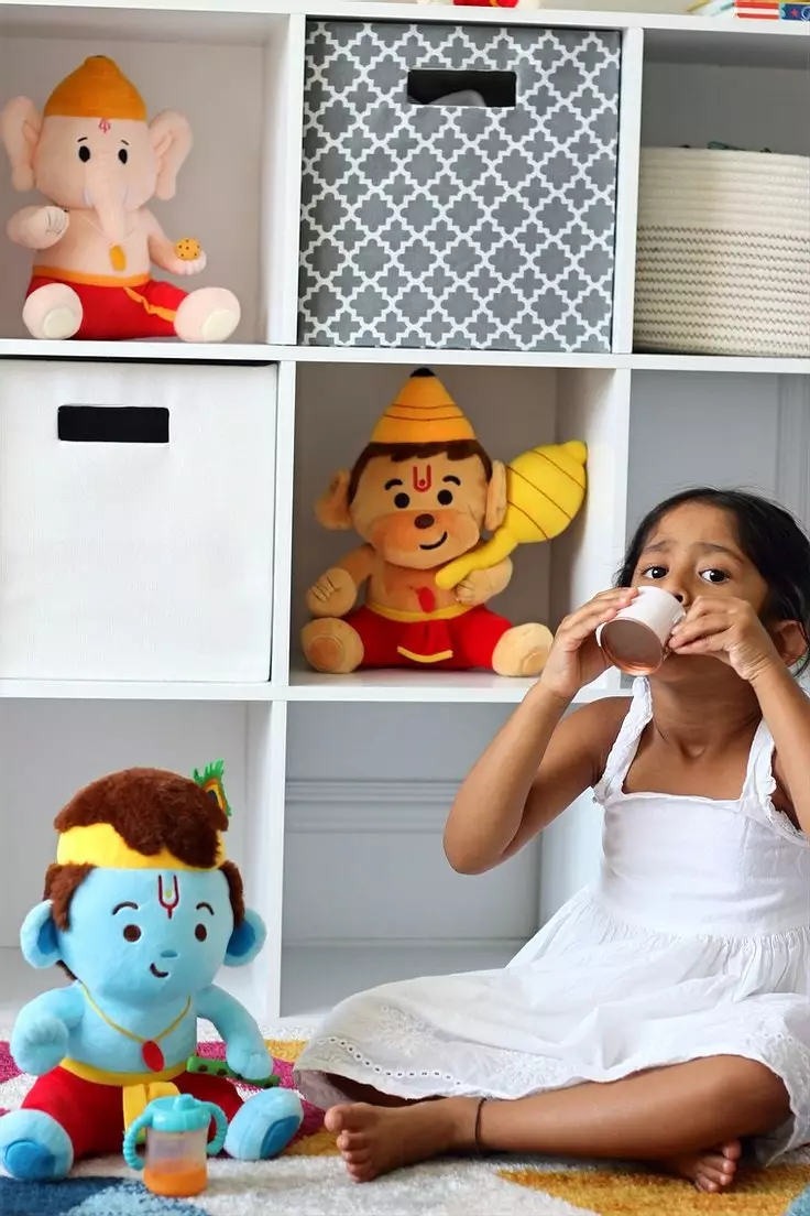 Een klein meisje drinkt thee voor een boekenplank met Modi Toys' pluche Baby Ganesh, Baby Hanum...