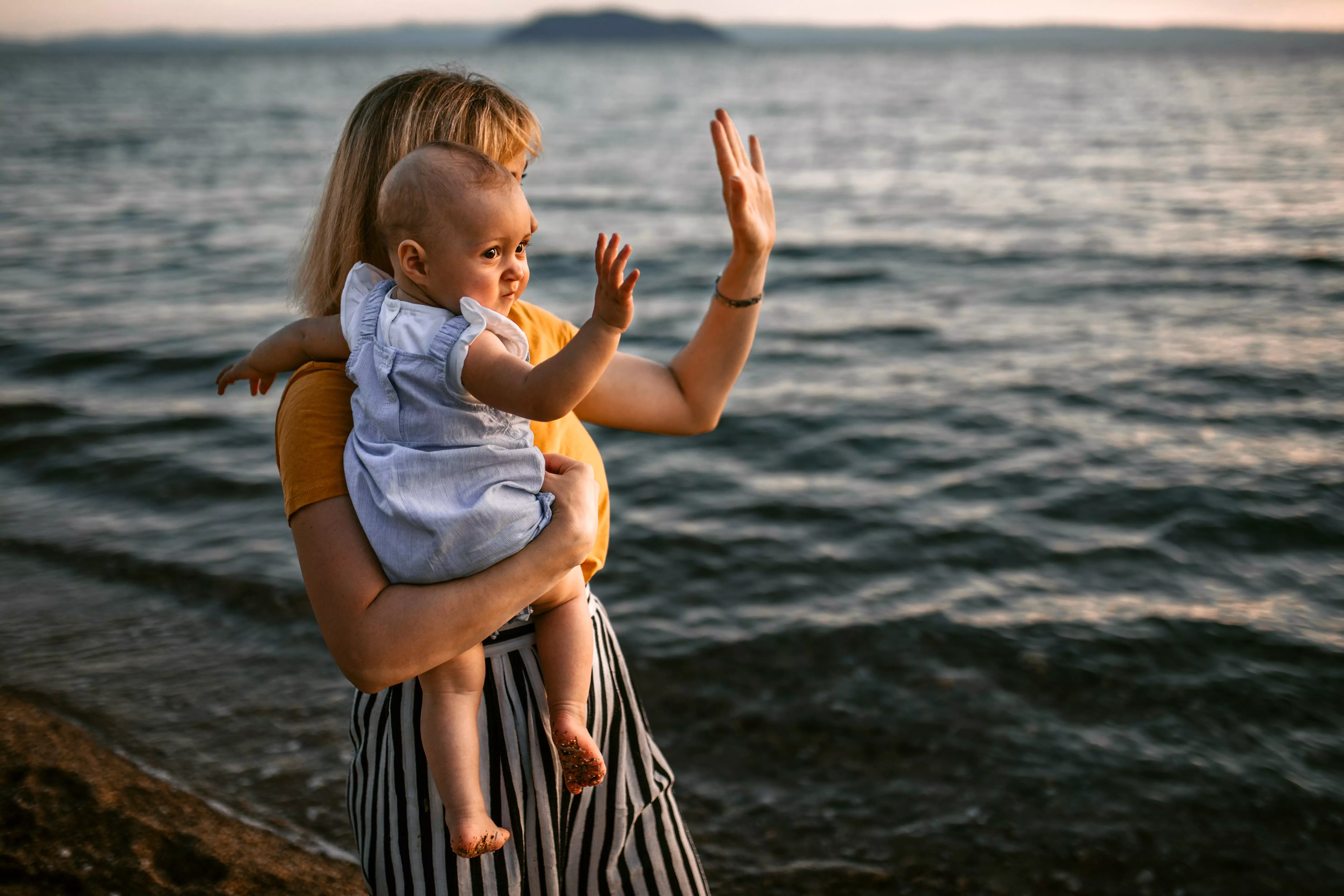 Mor og babyjente nyter kystvandring og vakker solnedgang på sandstranden og vinker til båtene